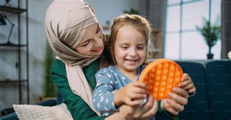 교육 어린이 게임 팝핏 안티 스트레스 감각 팝 그것 장난감 흥분하고 사랑스러운 어린 소녀 버튼으로 인기있는 안티 스트레스 게임을하고 히잡을 쓴 아랍 엄마와 소파에