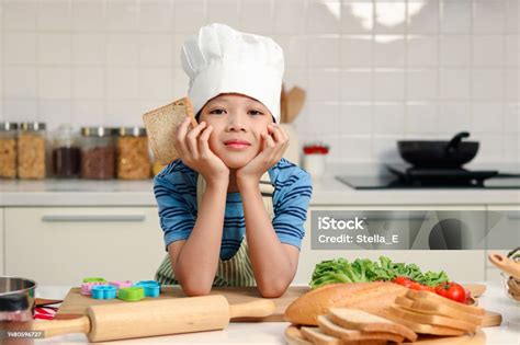 앞치마와 요리사 모자를 쓰고 행복한 미소를 짓고 있는 아시아 소년 아이의 초상화 부엌에서 건강한 식사를 요리하는 동안 손에 턱을 괴고 있는 귀여운 어린이 요리사는 혼자서