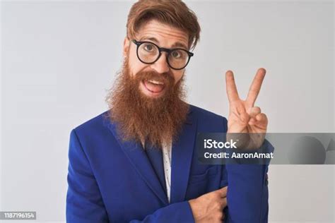 고립 된 흰색 배경 위에 양복과 안경을 착용한 젊은 빨간 머리 아일랜드 사업가가 승리의 표지판을하고있는 카메라에서 행복한 얼굴 윙크와 미소 2번 경영자에 대한 스톡 사진 및