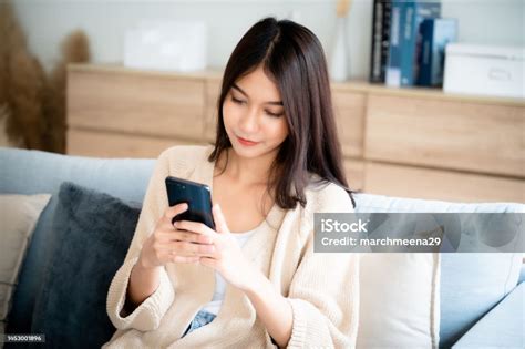 행복한 젊은 아시아 여성은 집에서 편안한 소파에서 휴식을 취하며 스마트 폰에서 문자 메시지를 보내고 웃는 소녀는 휴대 전화 채팅을 사용하고 가제트에서 무선 인터넷을 검색하고