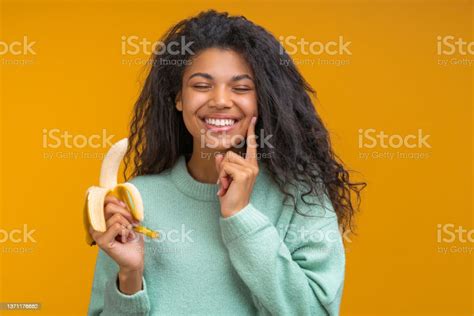 웃는 귀여운 옷을 입은 갈색 머리 소녀의 스튜디오 초상화는 그녀가 손에 들고있는 신선한 잘 익은 알약 바나나를 먹을 것입니다
