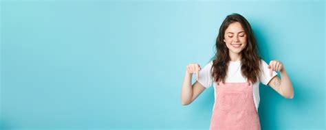 Симпатичная кавказская девушка в комбинезоне указывающая и смотрящая вниз радостно улыбаясь