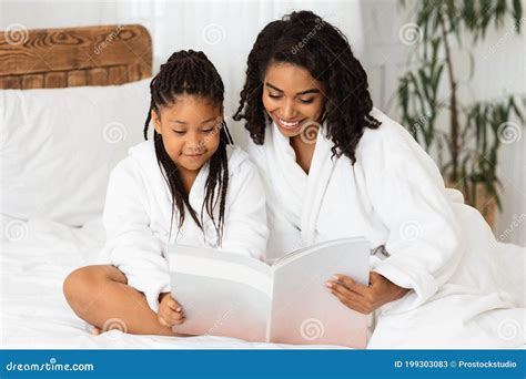 Feliz Madre E Hija Negras En Albornoces Sentados En La Revista De Lectura De La Cama Imagen De