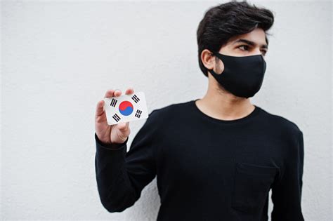 아시아 남자는 얼굴 마스크와 함께 모든 검은 색을 입고 흰색 배경에 고립 된 손에 한국 국기를 들고 코로나 바이러스 국가 개념