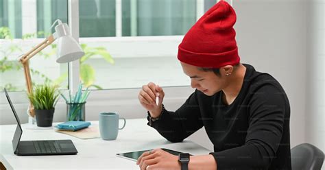 빨간 양모 모자를 쓴 젊은 남성 디자이너는 사무실에서 디지털 그래픽 태블릿과 스타일러스 펜을 가지고 책상을 작업하고 있다 사진
