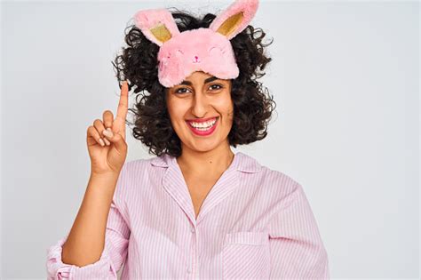 팔자마와 수면 마스크를 쓰고 고립된 흰색 배경을 통해 곱슬 머리를 한 아랍 여성은 자신감과 행복한 미소를 지으면서 손가락 번호 1을 가리키고 있습니다 가리키기에 대한 스톡