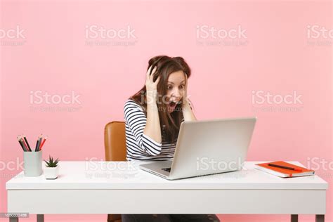 머리를 기대 며 비명 캐주얼 옷에 미친 여자 파스텔 핑크 배경에 고립 된 사무실에 앉아 있는 동안 노트북으로 프로젝트에서 작업