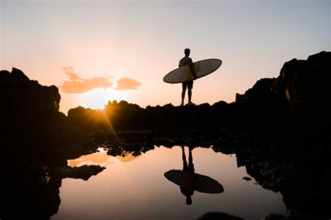 일몰에 남성 서퍼는 바다 해안의 절벽에 서핑 보드와 함께 서있다 파도를 기다리고 있습니다 여름 따뜻한 태양의 광선에 스포츠맨 남자의 실후트 서핑 캠프 경외감에 대한 스톡