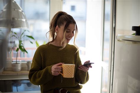 부엌에 서서 스마트 폰을 사용하는 동안 차 한 잔을 마시는 여성 Brand Name Online Messaging Platform에 대한 스톡 사진 및 기타 이미지