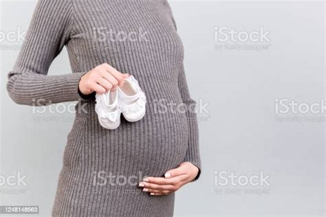 회색 배경에 임신 한 여성의 손에 아기 소녀를위한 신발의 클로즈업 미래의 어머니는 회색 드레스를 입고 있습니다 모성 개념 복사
