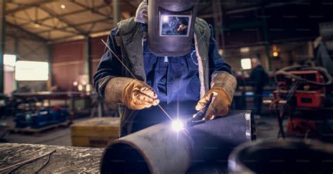 몇 안 되는 다른 작업자 앞에서 산업용 직물 작업장의 테이블에서 금속 조각 작업을 하는 제복을 입은 전문 마스크 보호 용접공 남자의 초상화 보기를 닫습니다 사진