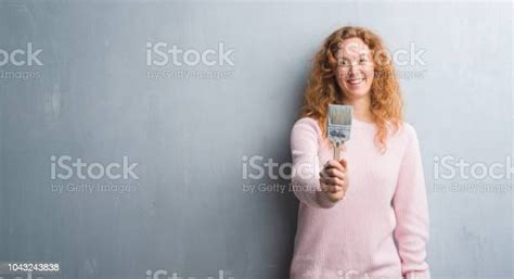 서 치아를 보여주는 자신감 미소로 웃 고 행복 한 얼굴로 페인트 붓을 들고 회색 그런 지 벽에 빨강 머리 젊은 여자 건강한 생활방식에 대한 스톡 사진 및 기타 이미지