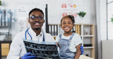 아프리카 의사가 작은 여성 환자를 껴안고 엑스레이 스캔을 손에 들고 있는 초상화 병원 사무실에 앉아 카메라를 향해 웃고 있는 귀여운 소녀와 함께 소아과 의사 사진