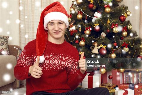 젊은 남자가 크리스마스 트리 근처 레트로 스웨터와 Sanat 모자 앉아 입고 새 해 이브 Ornated 전나무 나무입니다 겨울 휴가 사랑 개념입니다 12월 31일에 대한