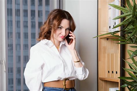 흰 블라우스를 입은 비즈니스 여성이 사무실 창문에 서서 휴대폰으로 이야기한다 40 44세에 대한 스톡 사진 및 기타 이미지 40 44세 40 49세 갈색 머리