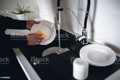 노란 스폰지와 흰색 접시를 세척 하는 남자의 손의 그림입니다 수돗물이 싱크대에 쏟아져 나오자 젊은 남자는 현대적인 부엌에 서있다