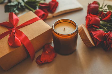 빨간 리본으로 묶인 선물 상자 옆에 놓인 나무 뚜껑이 있는 호박색 항아리 양초는 신선한 빨간 장미와 흩어진 꽃잎으로 둘러싸여 있습니다 펼쳐진 책은 아늑하고 낭만적인 느낌을