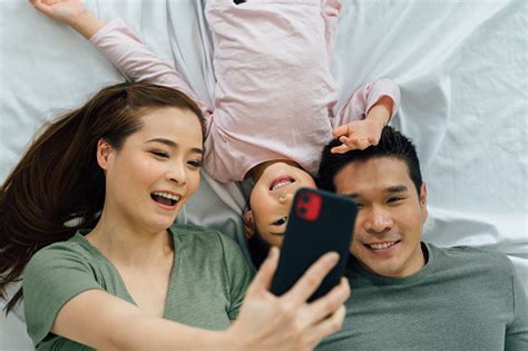 아시아의 어머니와 아버지는 스마트폰으로 사진을 찍는 침대에 귀여운 딸과 함께 누워 있습니다 3 명에 대한 스톡 사진 및 기타