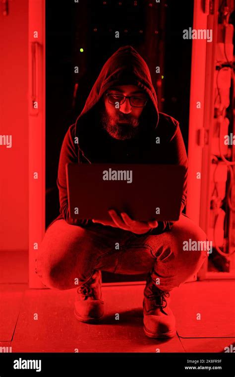 Computer Hacker With Laptop Crouching In Server Room With Red Light