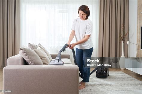 하얀 셔츠와 청바지를 입은 젊은 여성이 소파 아래 카펫을 청소하고 거실에 진공 청소기를 들고 공간을 복사합니다 집안일 청소 및
