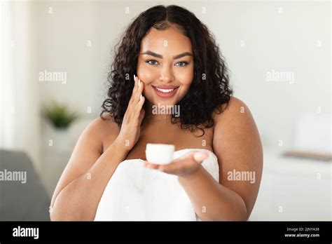 Happy Black Chubby Lady Using Face Cream At Home Looking At Camera And Showing Cream Jar