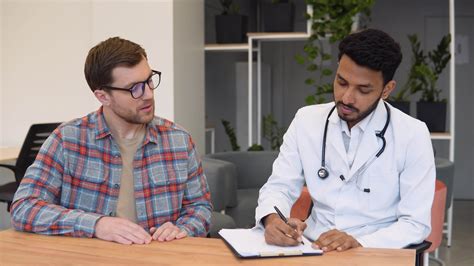 Indian Male Doctor Consulting Patient Filling Form At Consultation