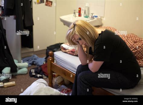 Young Teenage Woman Sitting On Bed In Messy Hmo Bedsit Bedroom Holding