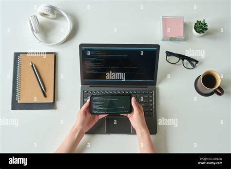 Above View Of Programmer Connecting Smartphone With Codes With Laptop At Her Workplace Stock