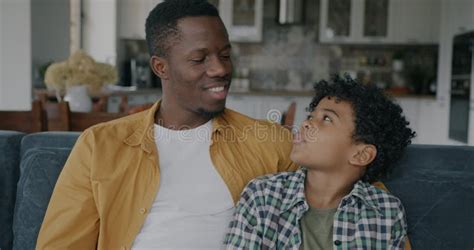 Father And Son Talking Smiling Then Looking At Camera And Smiling Sitting On Couch In Apartment