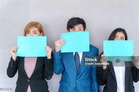 젊은 사업가 그룹과 사업가그룹은 회색 배경에 서 있는 무언가를 바라보면서 녹색 빈 종이를 들고 얼굴의 절반을 덮는 양복을 입고 있습니다 20 24세에 대한 스톡 사진 및