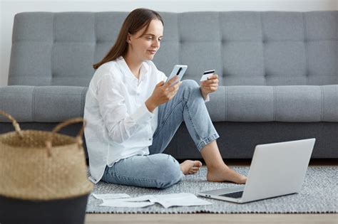 소파 근처 바닥에 앉아 흰색 셔츠와 청바지를 입고 노트북과 전화를 사용하여 화면을 보고 신용 카드를 들고 있는 백인 여성의 수평