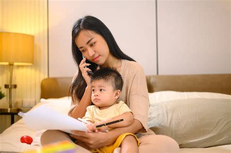 Busy Working Mom Talking On Mobile Phone And Holding Her Baby Son Sitting On Bed Stock Photo