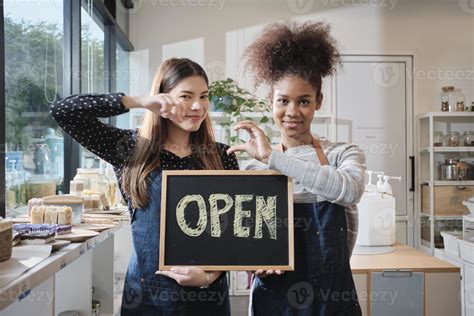 Two Young Female Shopkeepers Show Open Sign Board With Cheerful Smiles