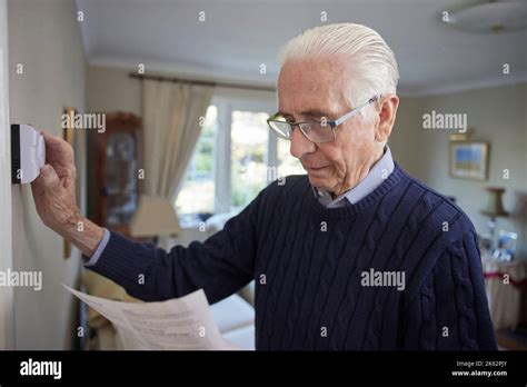 Worried Senior Man With Bill Turning Down Central Heating Thermostat At Home In Energy Crisis