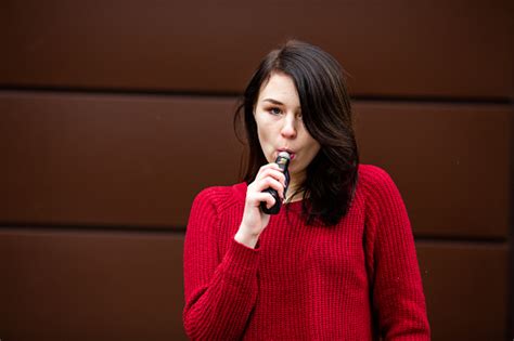 Vape 십대 겨울에 거리에서 현대 갈색 배경 맞은 편에 전자 담배를 흡연 빨간 스웨터에 젊은 예쁜 흰색 백인 소녀 나쁜 습관