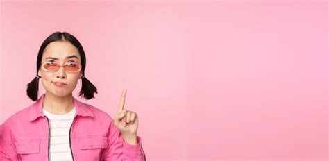 Premium Photo Hesitant Asian Girl Wears Sunglasses Frowns And Points