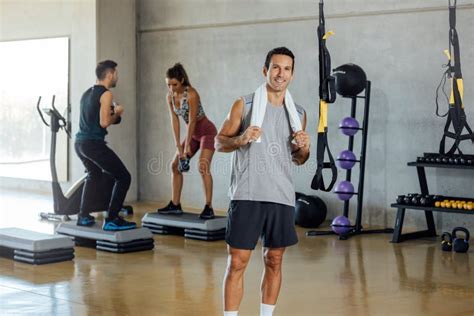 Happy Healthy Man Standing In Gym While People In The Back Exercises