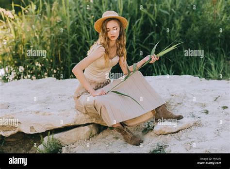 Attractive Blonde Girl In Elegant Dress And Straw Hat Sitting On Ground