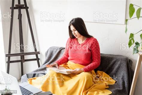 캐주얼 한 옷과 격자 무늬에 쾌활한 미소 소파에 앉아 집에서 책을 읽고 즐기는 예쁜 젊은 여성 가정 생활에 대한 스톡 사진 및
