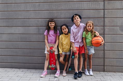 도시에서 함께 포즈를 취하고 콘크리트 벽 앞에 서서 스케이트 보드를 들고 카메라를 보는 행복한 친구들 아이에 대한 스톡 사진 및