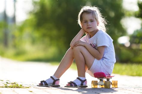 흰색 반바지와 티셔츠에 꽤 귀여운 작은 금발 소녀는 분홍색 스케이트 보드에 앉아 밝은 흐린 여름 배경에 카메라에 미소 어린이 활동과 활동적인 라이프 스타일 개념 감정에 대한
