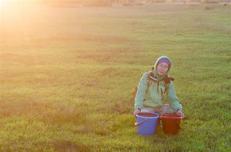 フードと帽子をかぶったヤクートアジアの少女と日没の太賀のスプルースの森に向かって緑の野原に座り北部の野生のクランベリーのバケツを2つ集めた