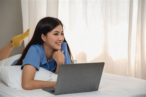 아름다운 얼굴 긴 머리 파란 셔츠를 입은 젊은 아시아 여성이 집에서 하얀 침실의 침대에 누워 커피 한 잔과 노트북으로 화상