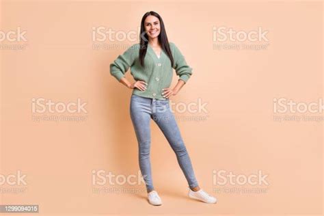 긍정적 인 행복한 여성의 전체 길이 사진은 베이지 색 배경에 고립 된 그린 카디건 청바지를 입고 손 허리를 잡고 카디건 웃옷에