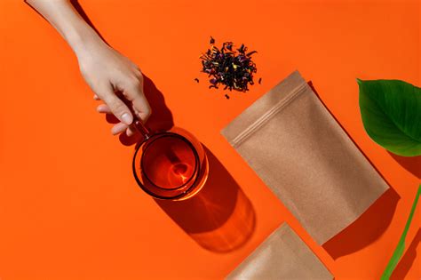 차 한 잔과 함께 차와 여성 손을위한 갈색 판지 포장 오렌지색 배경에 차 브랜딩 및 포장의 모형 가방에 대한 스톡 사진 및 기타