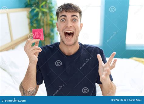Young Hispanic Man Sitting On The Bed Holding Condom Celebrating