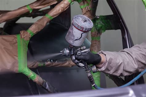 보호 작업 바지와 마스크에 남자는 그의 손에 스프레이 병을 보유 하 고 차량 복구 워크샵에서 수리 하는 동안 사고 후 차체의 프레임에 검은 페인트 스프레이 교통사고 충돌